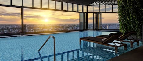 Piscina infinita no terraço com vista para a cidade ao pôr do sol, cadeiras de lounge de madeira e guarda-corpo de vidro.
