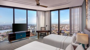 Quarto de hotel moderno com uma cama grande, uma TV em um móvel de madeira, uma mesinha e vista para a paisagem urbana através de grandes janelas.