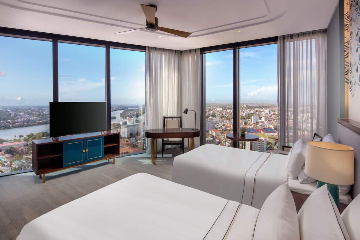Quarto de hotel moderno com uma cama grande, uma TV em um móvel de madeira, uma mesinha e vista para a paisagem urbana através de grandes janelas.