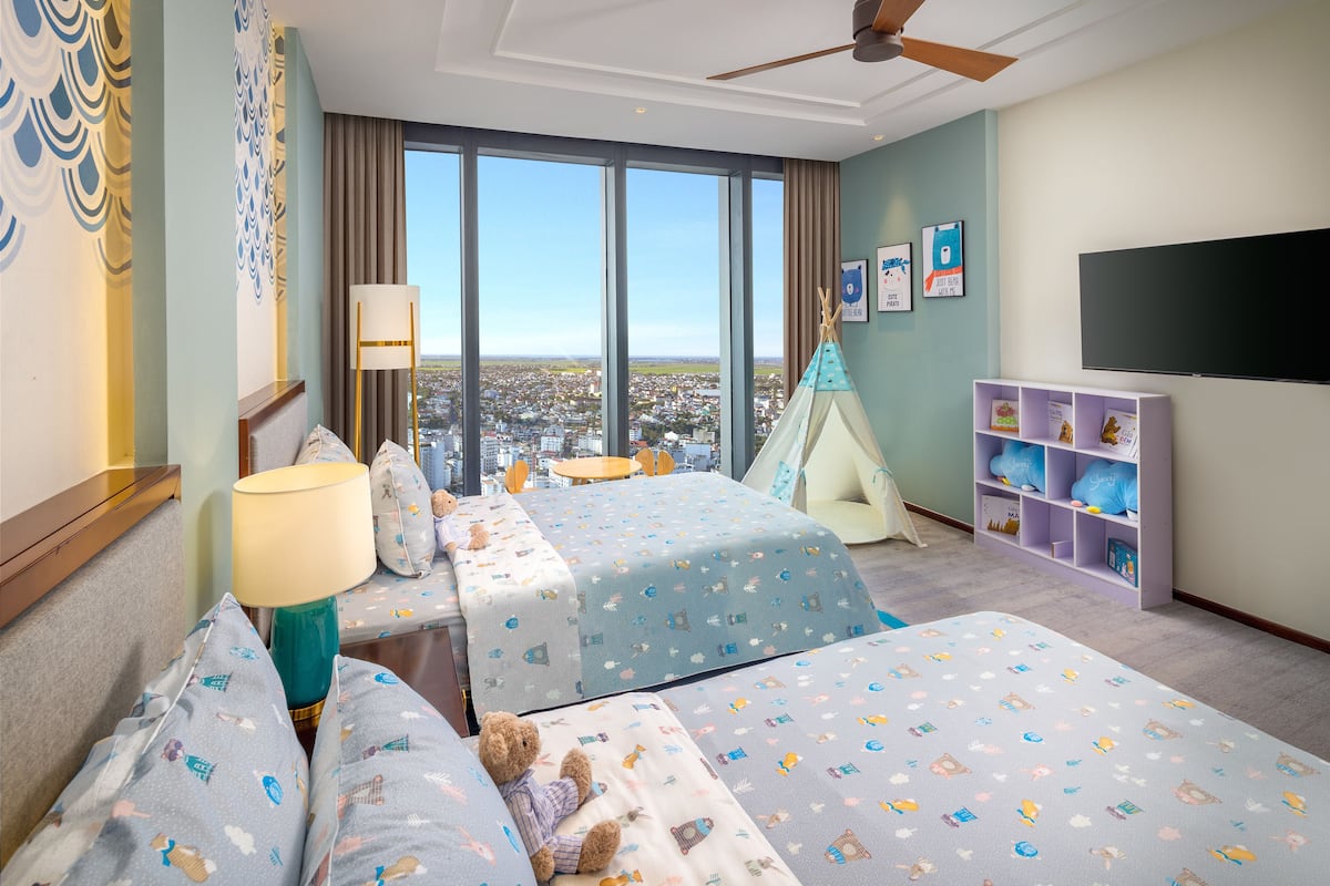 Quarto de hotel com duas camas, uma janela grande com vista para a cidade, um ventilador de teto e uma estante de livros.