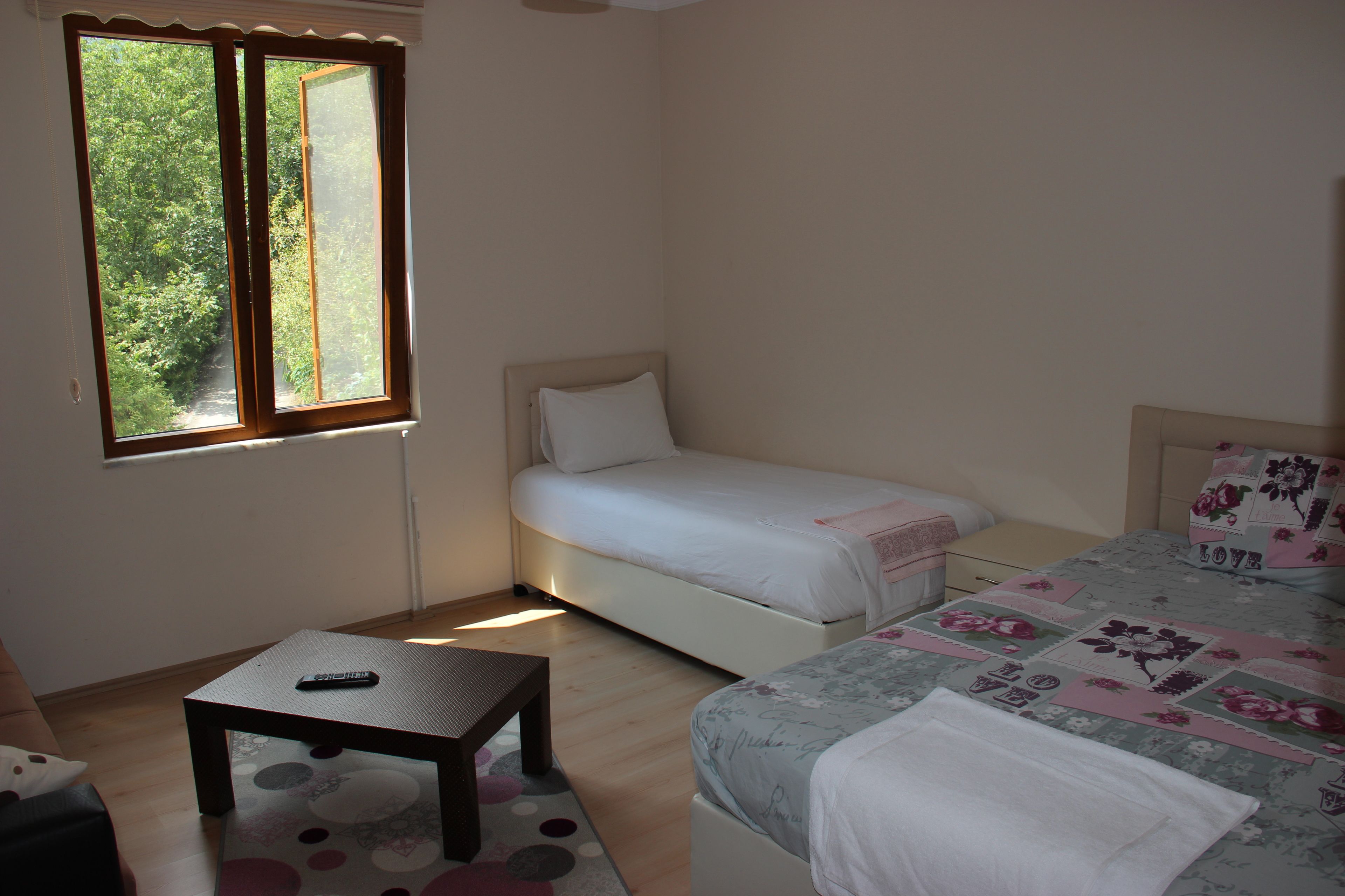Quarto pequeno e bem iluminado, com uma cama de solteiro, mesa de cabeceira, janela com vista para vegetação e uma pequena mesa.