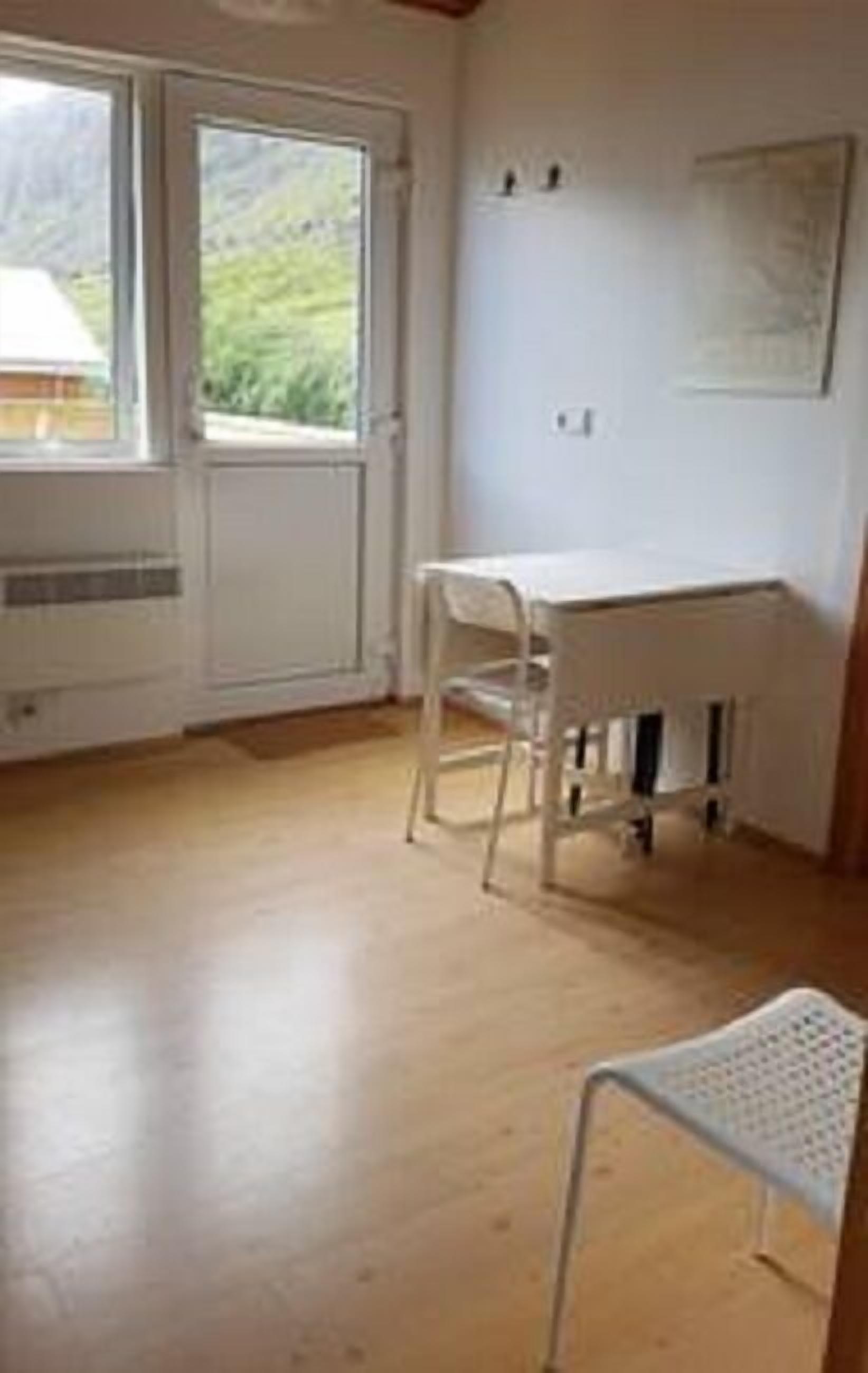 Quarto com piso de madeira, uma mesa branca e uma cadeira branca. Há uma janela com vista para o verde lá fora.
