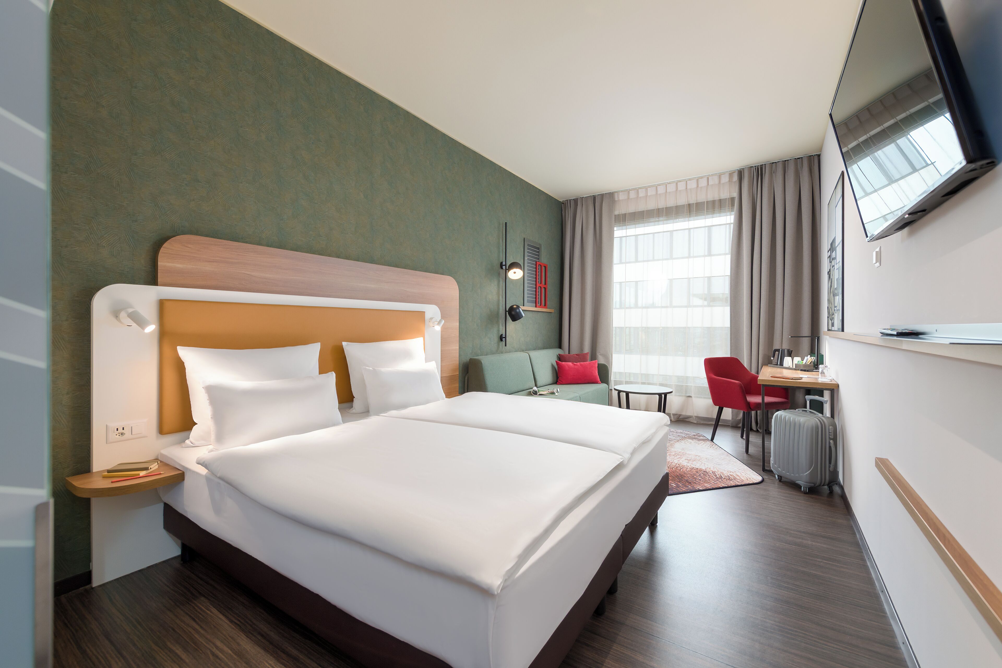 Quarto de hotel com uma cama grande, uma escrivaninha, uma cadeira, uma televisão e uma janela com cortinas.