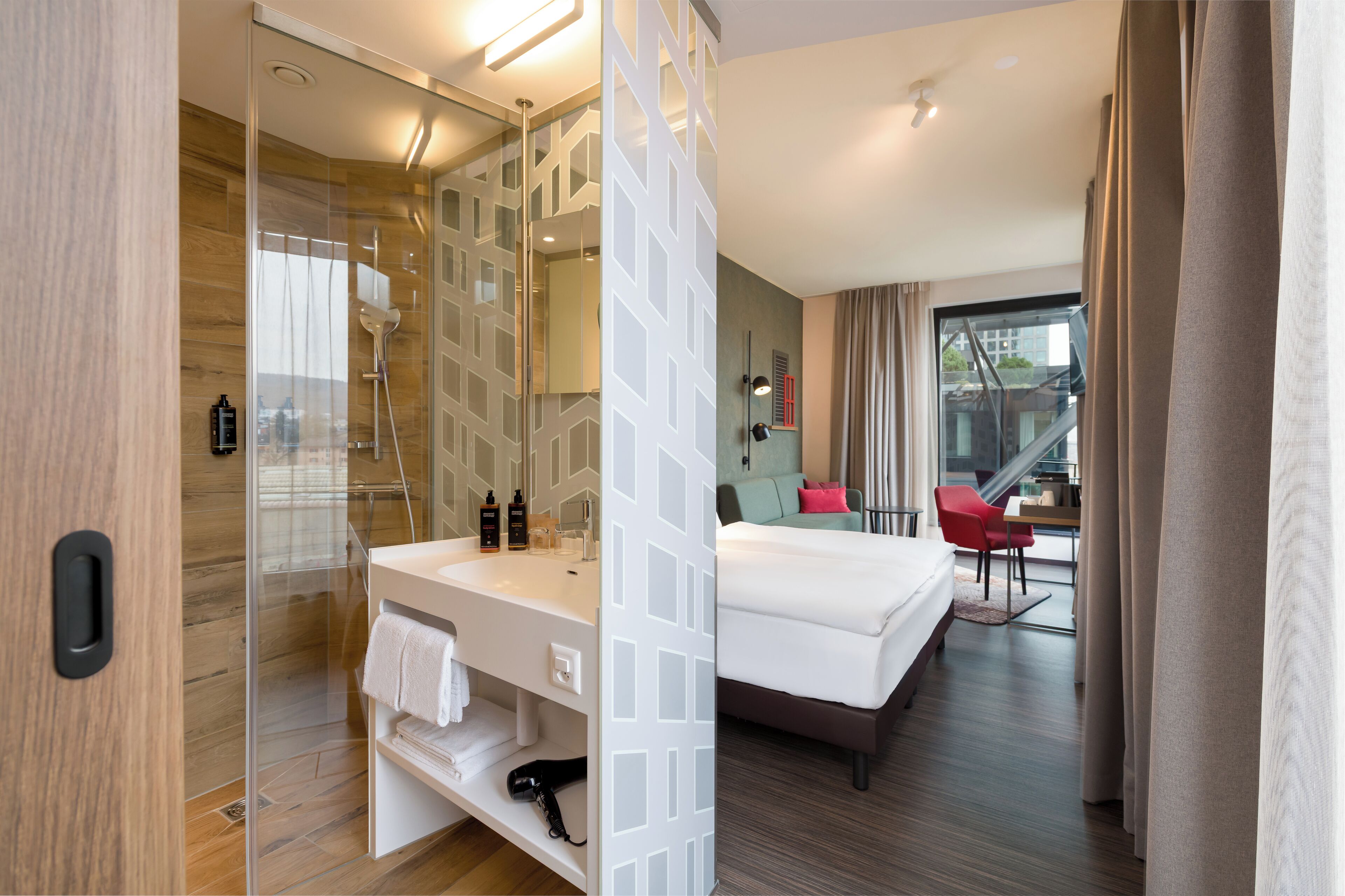 Quarto de hotel moderno com chuveiro em cabine de vidro, uma cama grande, uma escrivaninha e uma cadeira vermelha.