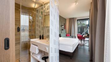 Quarto de hotel moderno com chuveiro em cabine de vidro, uma cama grande, uma escrivaninha e uma cadeira vermelha.