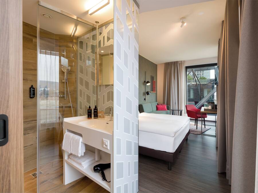 Quarto de hotel moderno com chuveiro em cabine de vidro, uma cama grande, uma escrivaninha e uma cadeira vermelha.