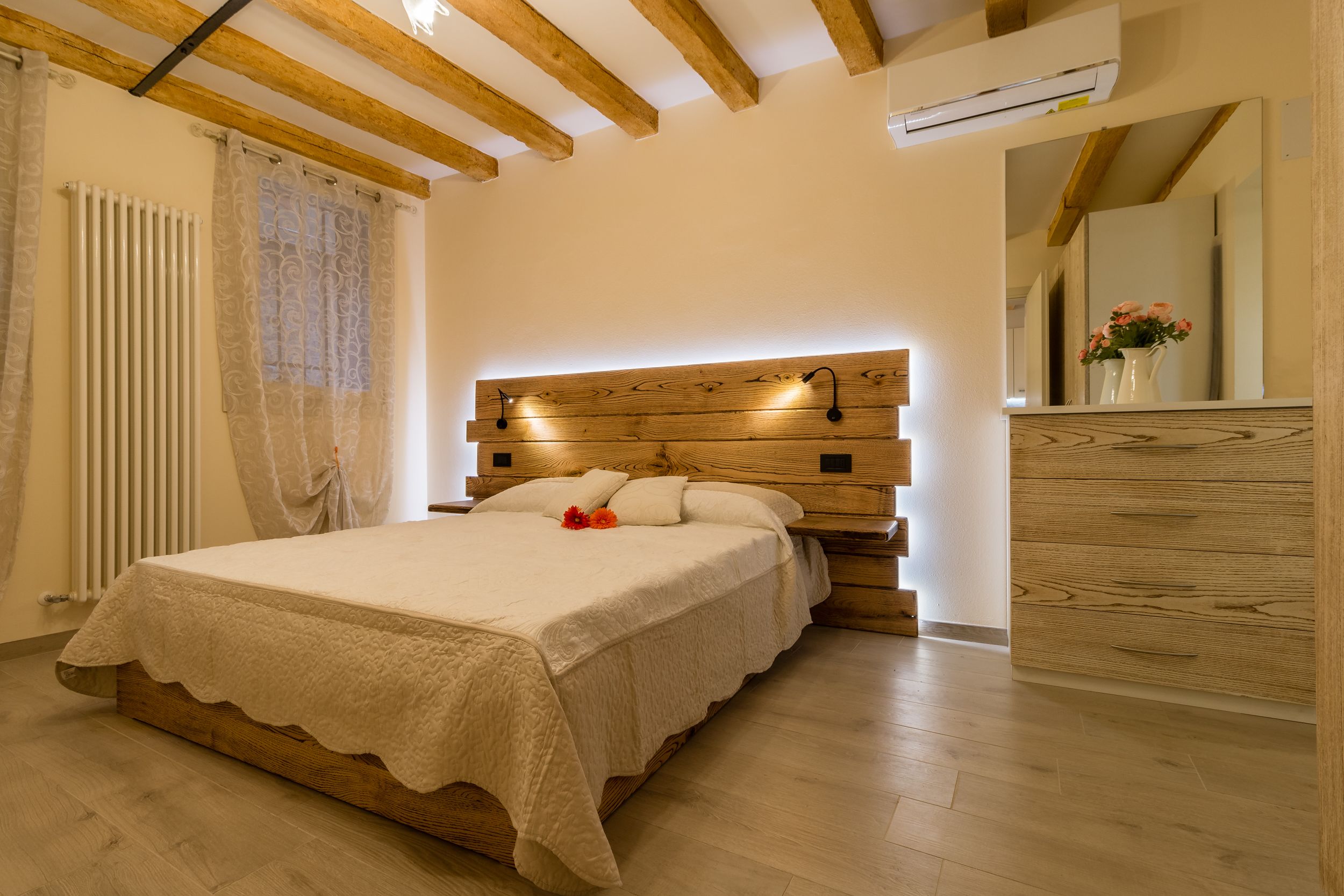 Quarto com cabeceira de madeira, cama com colcha branca, mesa de cabeceira com um vaso de flores e cômoda de madeira.