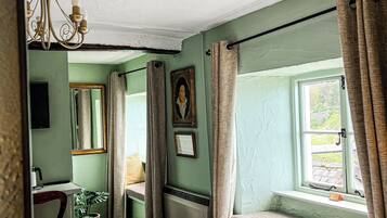 Um quarto com cama, janela com cortinas, um espelho refletindo o quarto, radiador e um lustre.