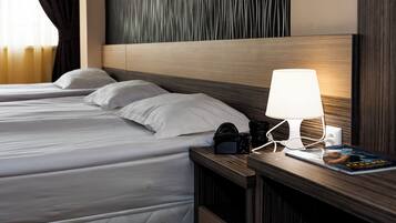 Quarto de hotel com duas camas, cabeceira de madeira, mesa de cabeceira com abajur, uma câmera e uma revista.