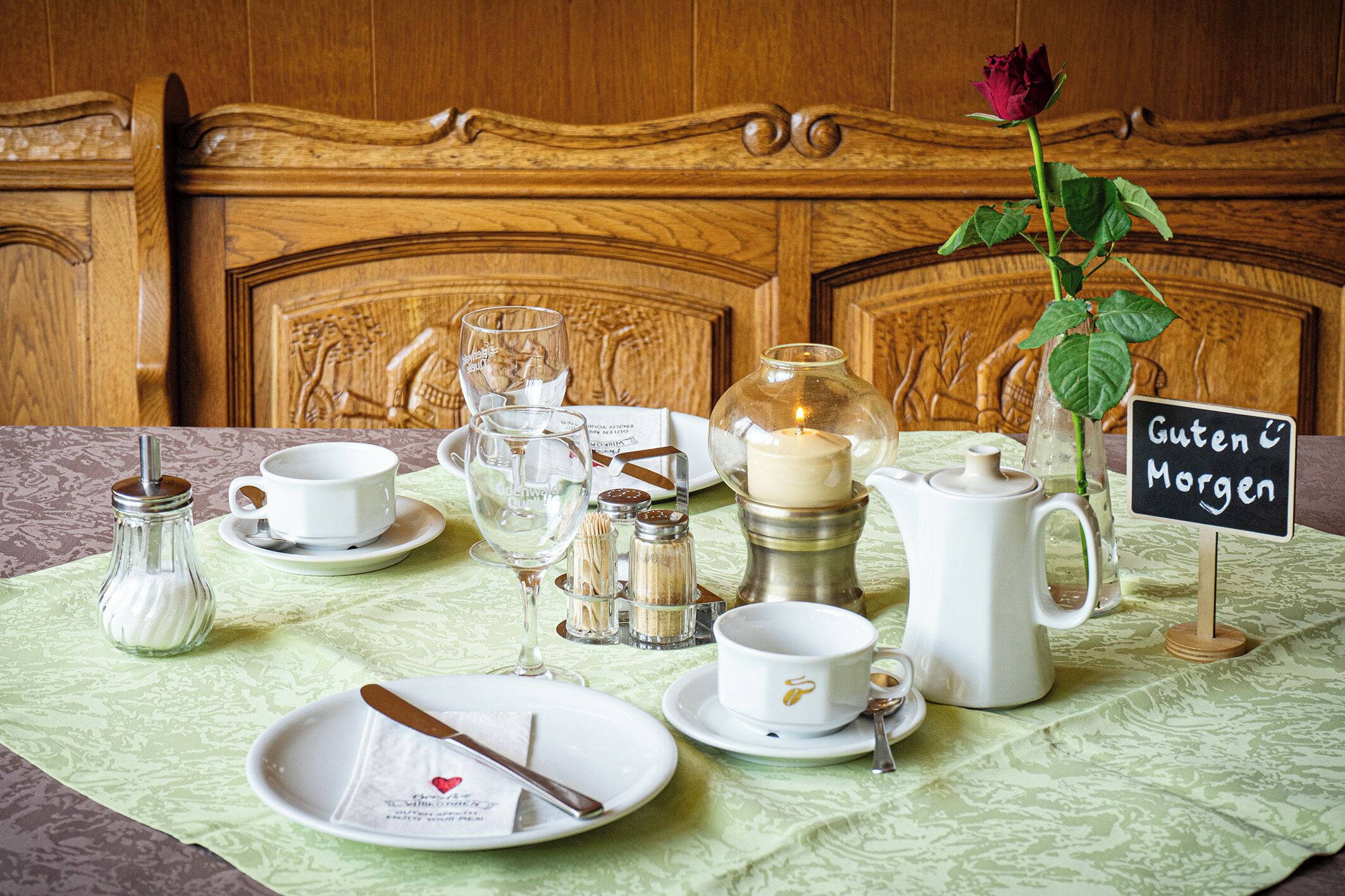 Disposição para o café da manhã com uma mesa posta para dois, incluindo pratos, xícaras, um bule e uma placa com a inscrição 'Guten Morgen'.