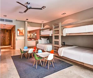 Quarto de hotel com beliches, área de refeições com cadeiras, ventoinha no teto e cozinha compacta.
