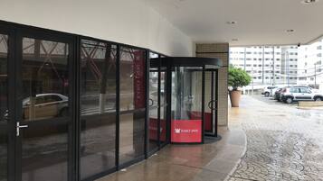 Entrada de um hotel com porta giratória, portas de vidro e calçada de azulejos.