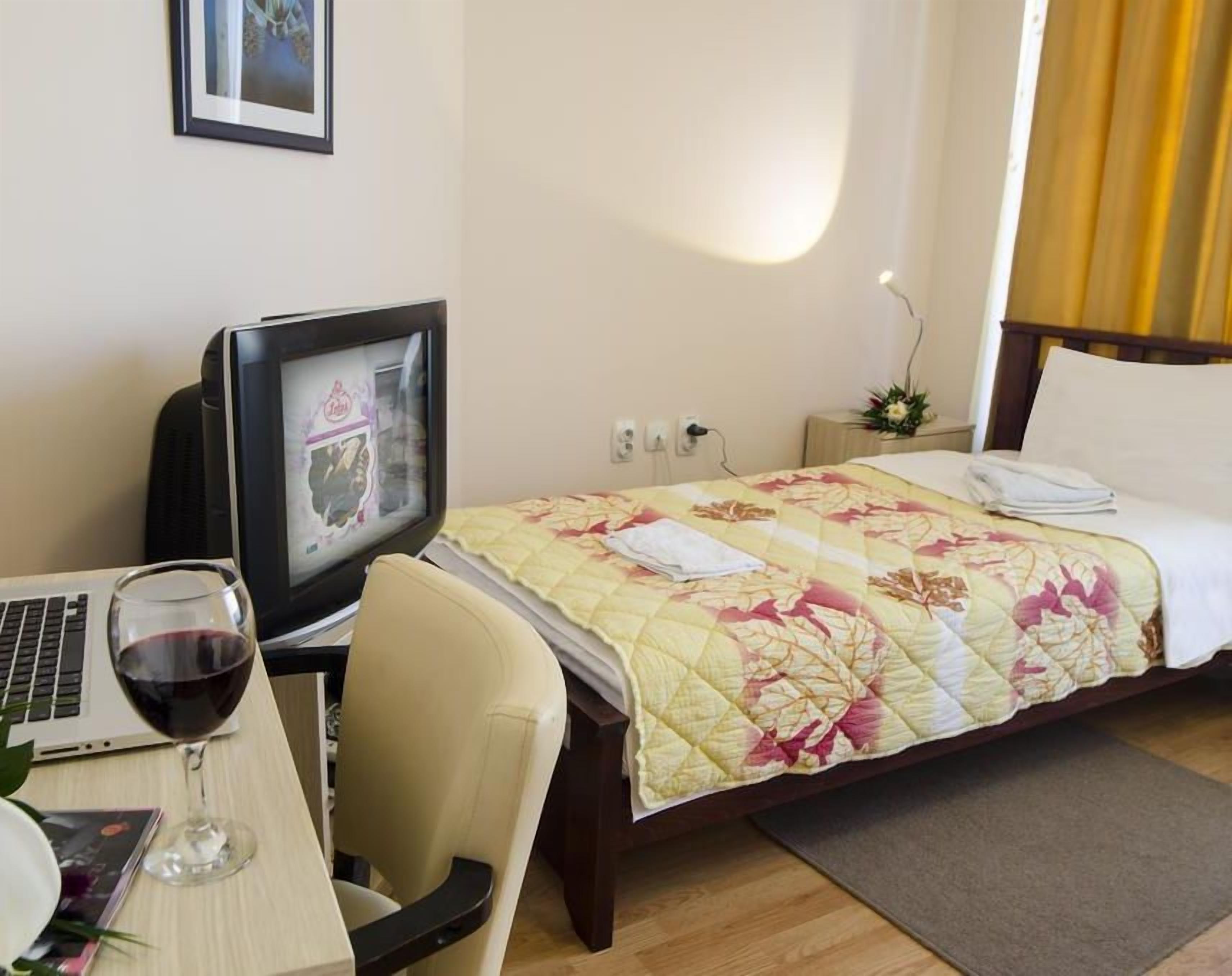 Quarto de hotel com cama, escrivaninha com um laptop e um copo de vinho, televisão e mesa de cabeceira com uma flor.
