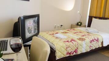 Quarto de hotel com cama, escrivaninha com um laptop e um copo de vinho, televisão e mesa de cabeceira com uma flor.