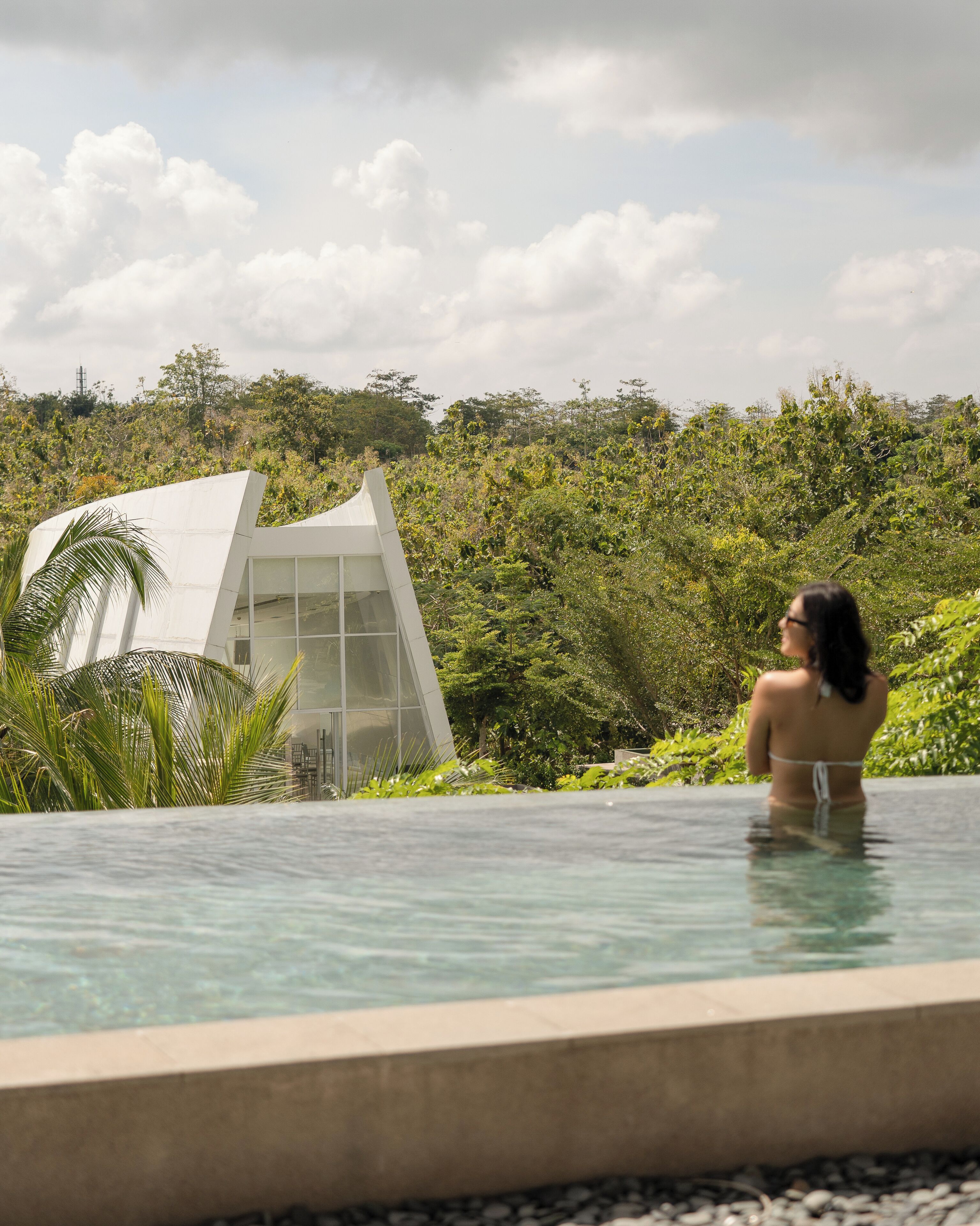 Mulher de biquíni branco em piscina transparente, com um edifício moderno branco e vegetação exuberante ao fundo.