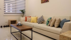 Sala de estar com sofá, almofadas, mesa de centro, planta e uma pintura abstrata colorida na parede.
