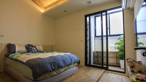 Quarto com cama, piso de madeira, porta de vidro deslizante que dá para uma varanda e uma mesinha com um brinquedo de pelúcia.