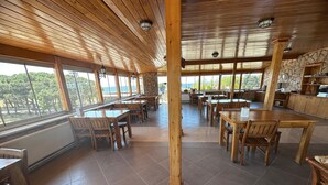 Uma sala de jantar ampla com mesas e cadeiras de madeira, grandes janelas com vista para o exterior e uma parede de destaque em pedra.