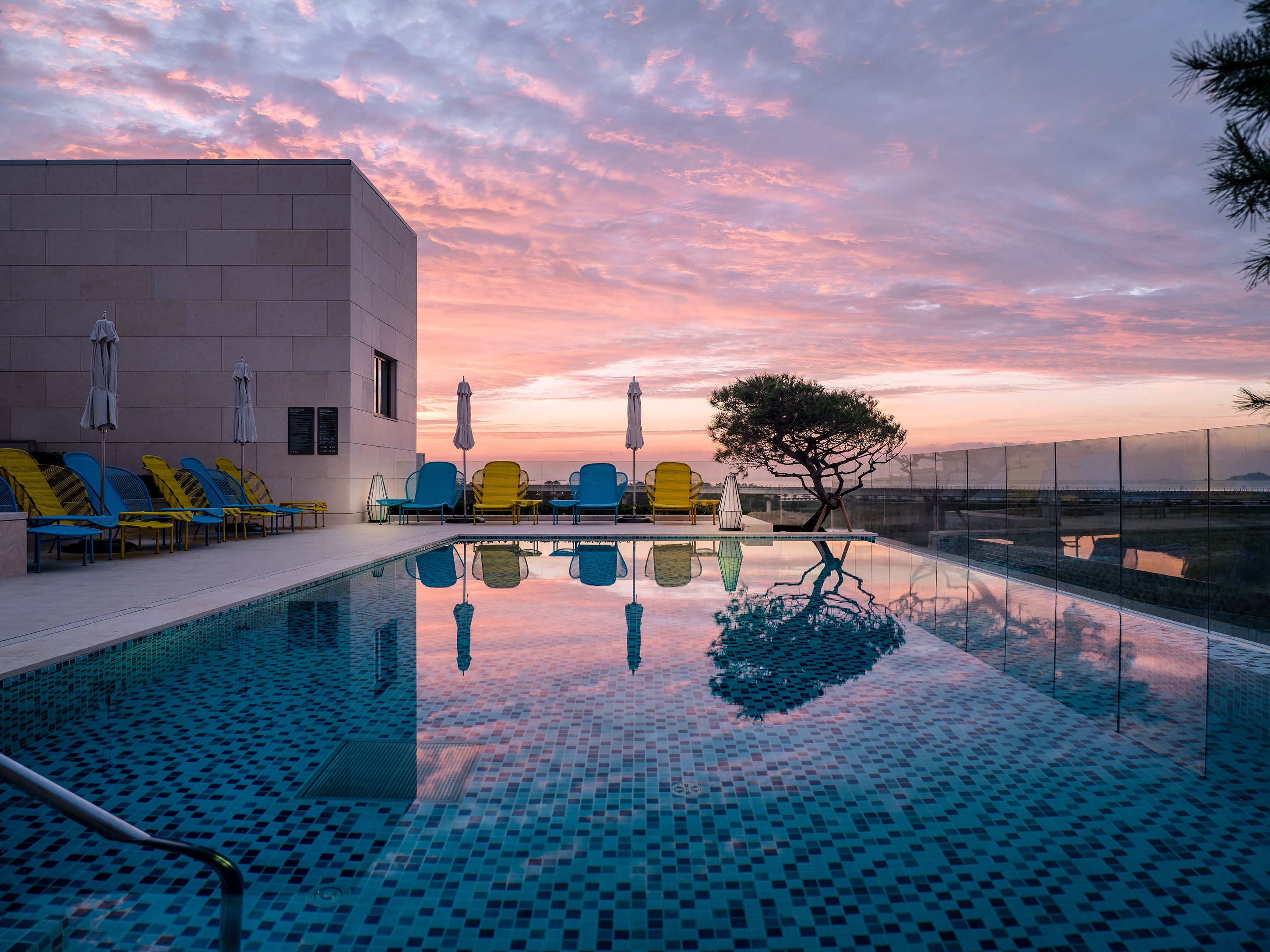 Uma piscina no terraço com cadeiras de descanso azuis e amarelas, uma árvore e vista para a cidade ao pôr do sol.