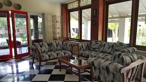 Um chão xadrez, um sofá e uma mesa de centro de vidro numa sala com janelas amplas e uma porta.