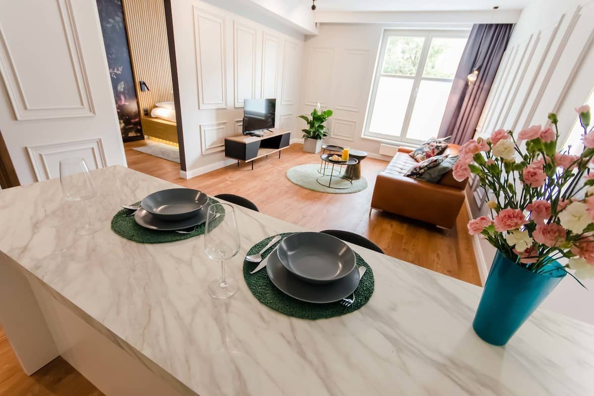 Cozinha moderna com bancadas de mármore, sala de jantar com mesa posta para dois, um sofá e uma televisão ao fundo.
