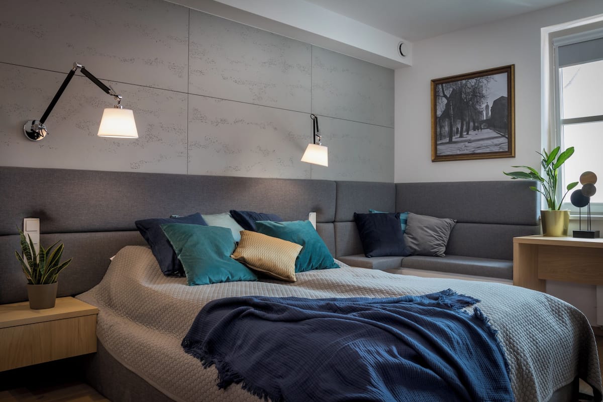 Quarto moderno com uma cama grande, duas luminárias de cabeceira, um sofá cinzento, um quadro na parede e um vaso com planta na mesinha de apoio.
