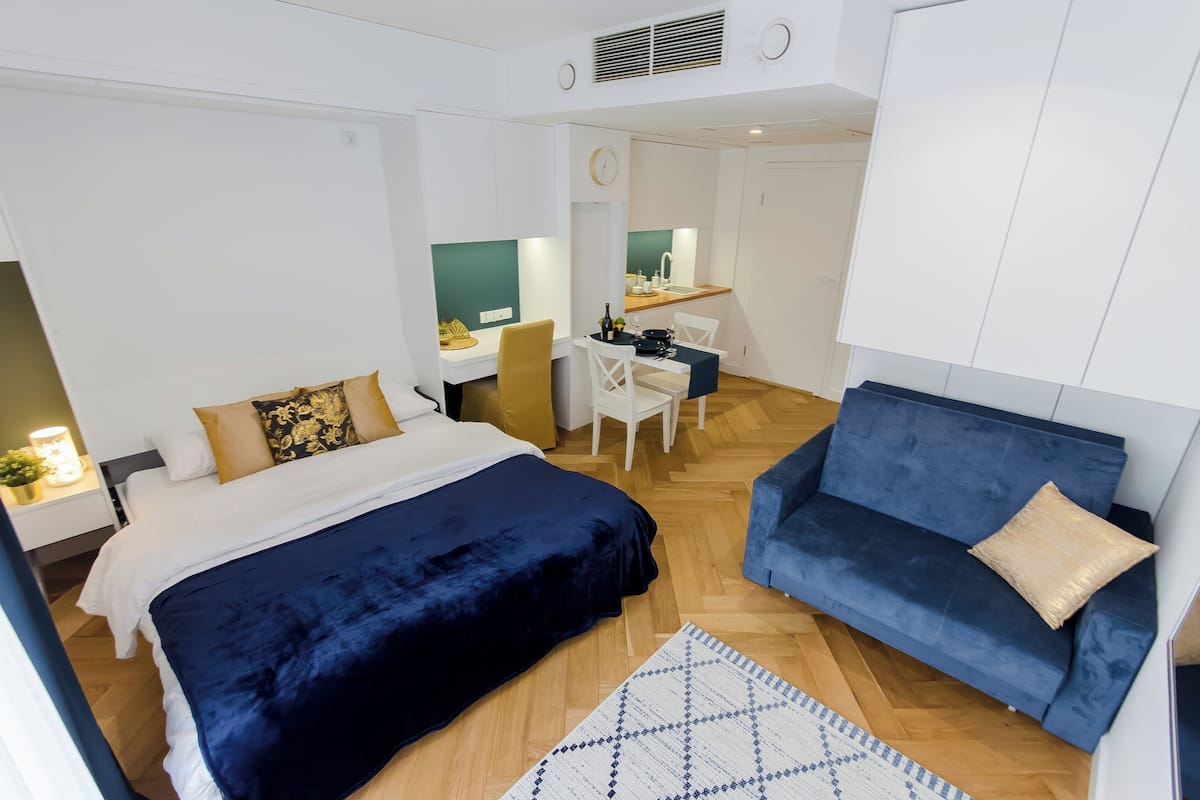 Quarto moderno com cama, sofá azul, escrivaninha com cadeira e pequeno espaço de cozinha.