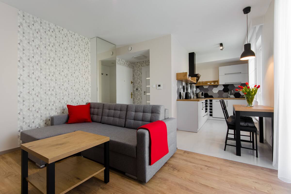 Uma sala de estar moderna com um sofá cinzento, uma mesa de centro de madeira e um cobertor vermelho. Uma área de cozinha é visível ao fundo, com uma mesa de jantar e cadeiras.