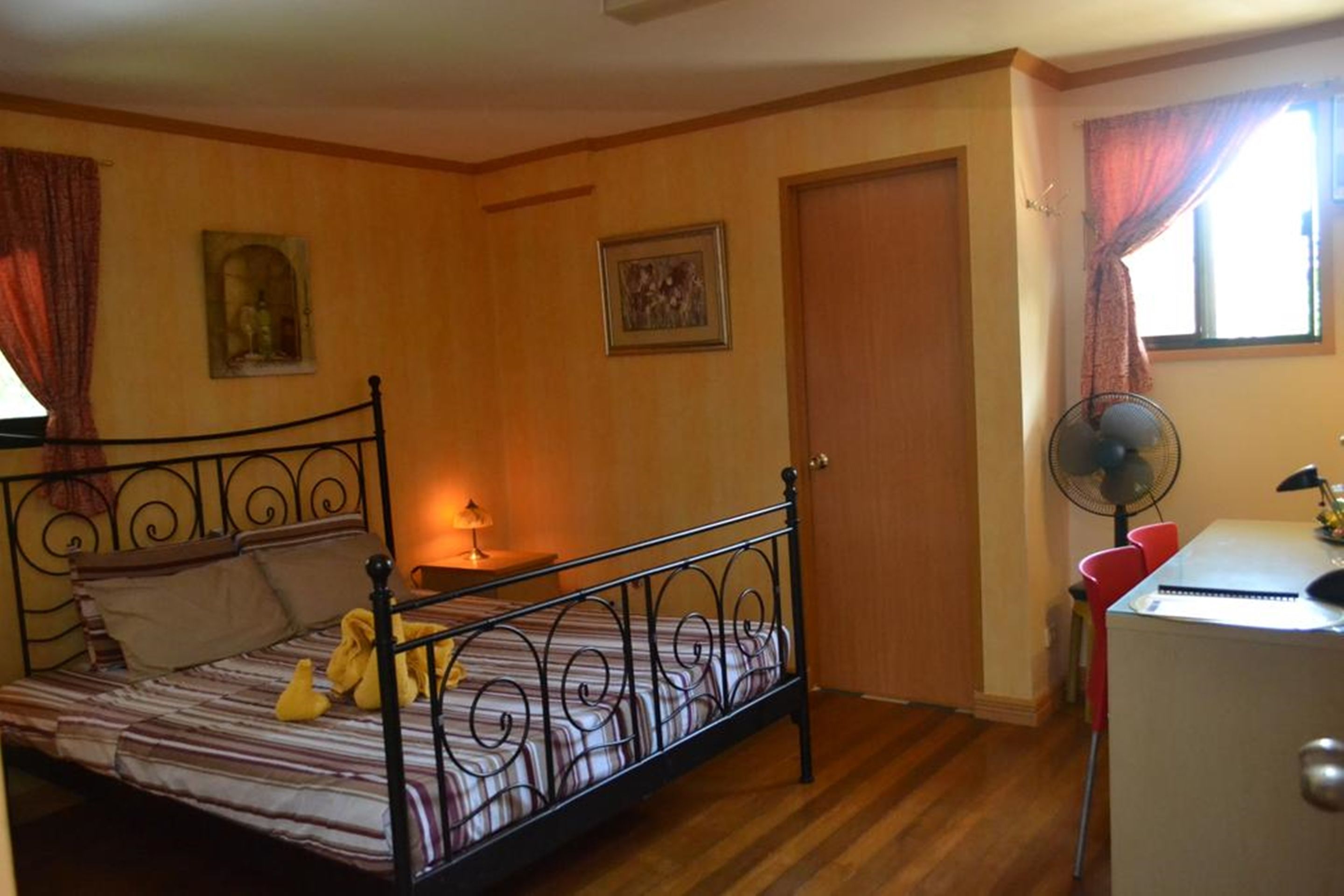 Quarto com cama, escrivaninha, cadeira, ventilador e duas pinturas na parede.