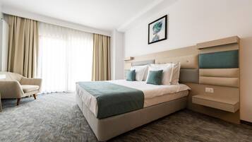 Quarto de hotel com uma cama grande, uma cadeira, cabeceira com compartimentos de armazenamento e um quadro na parede.
