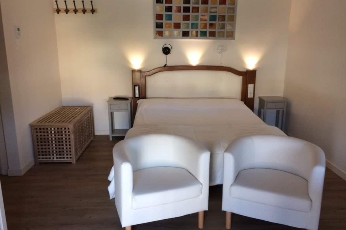 Quarto com cama, duas cadeiras brancas, cabeceira de madeira, mesas de cabeceira, luminária na parede e cesto de roupa.