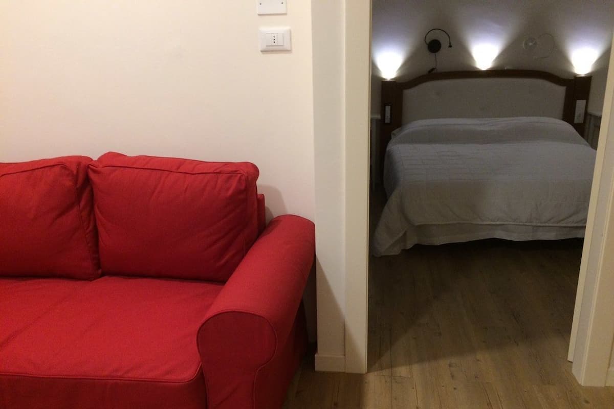 Um sofá vermelho em um quarto com piso de madeira e uma cama com colcha branca.