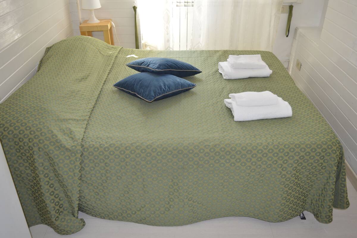 Cama bem arrumada com roupa de cama verde, dois travesseiros azuis e toalhas brancas dobradas.