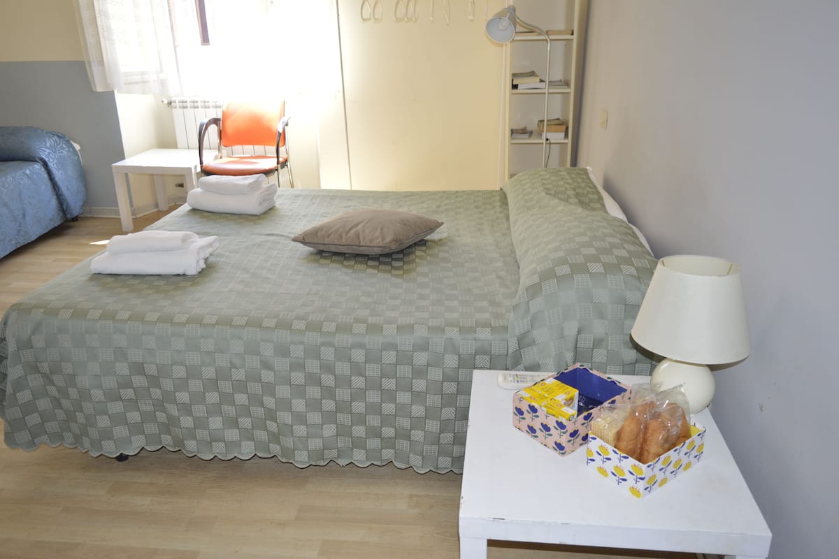 Cama bem arrumada com colcha xadrez, um travesseiro e toalhas dobradas. Há uma mesinha de cabeceira com uma luminária e uma cesta com pão e outros itens. Uma cadeira e uma estante com livros são visíveis ao fundo.
