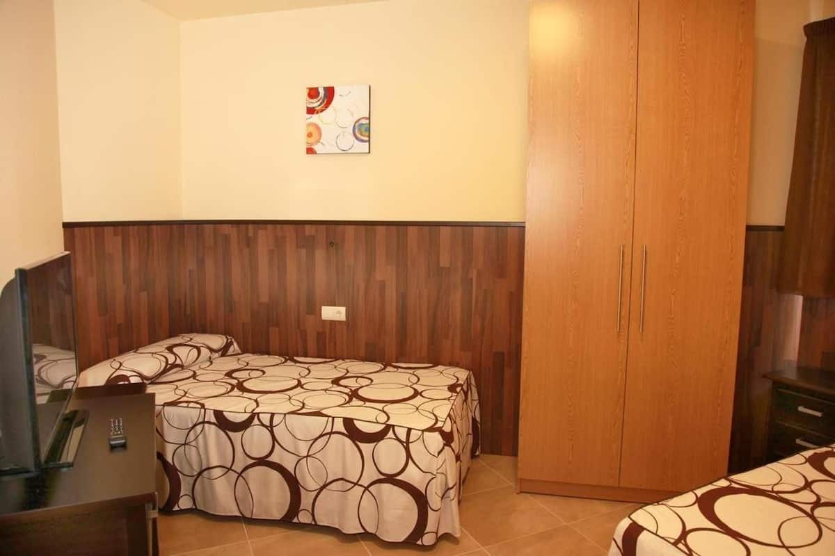 Quarto de hotel com duas camas, um guarda-roupa de madeira e uma secretária pequena com telefone.
