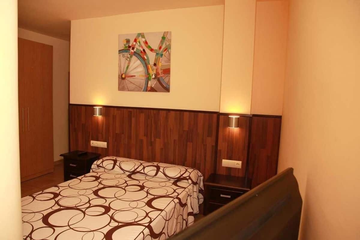 Quarto de hotel com cabeceira de madeira, cama com colcha estampada em padrão circular, mesa de cabeceira, pintura pendurada na parede e guarda-roupa de madeira.