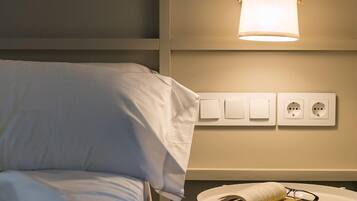 Cama bem arrumada com lençóis brancos, mesa de cabeceira com abajur e tomada na parede.