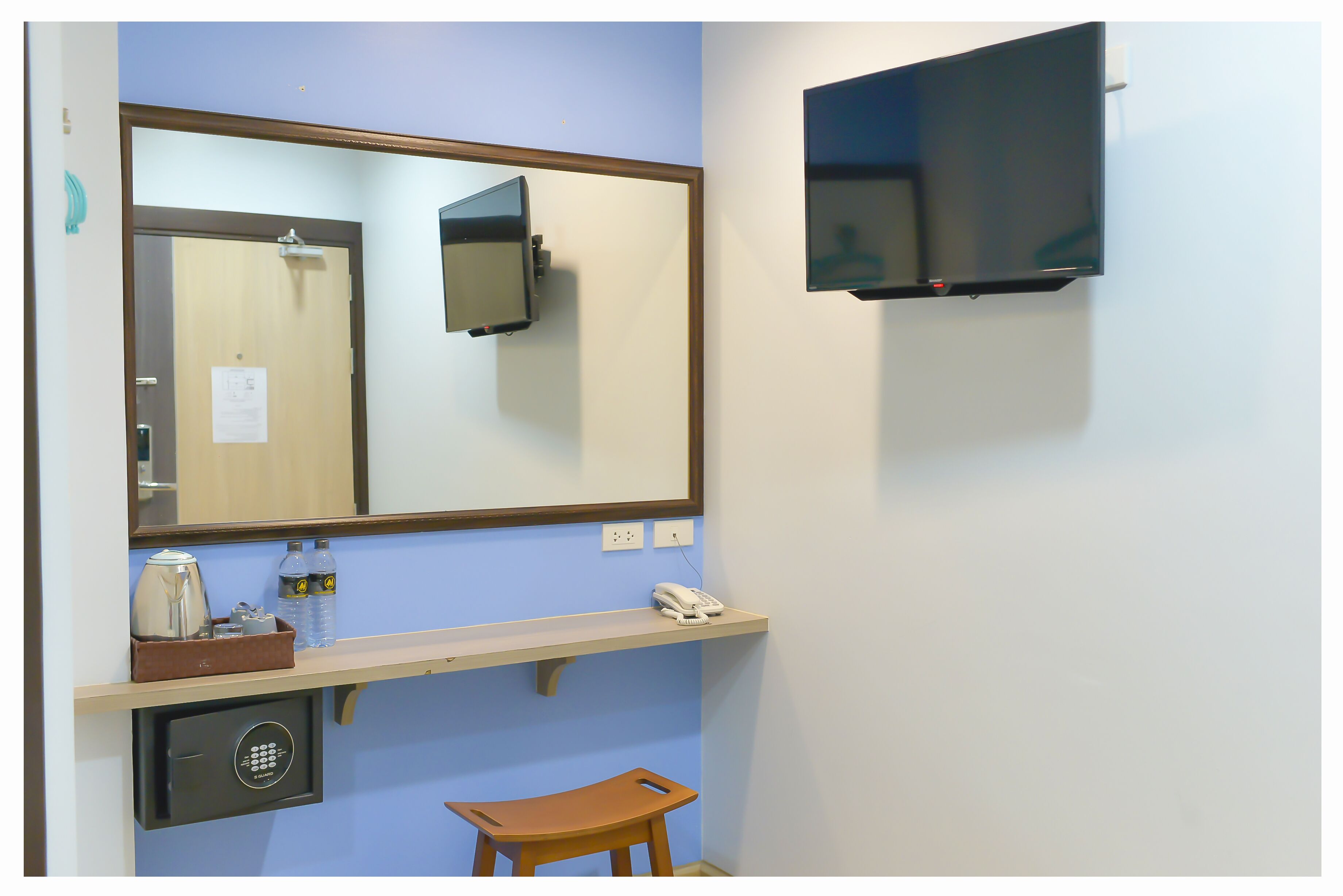 Quarto de hotel com um espelho grande, televisão de tela plana na parede, um banquinho de madeira, frigobar e telefone.