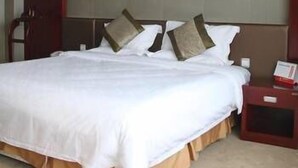 Uma cama bem arrumada com lençóis brancos e duas almofadas, uma mesa de cabeceira de madeira com um livro e um telefone, e uma parede lisa com uma cabeceira de madeira.
