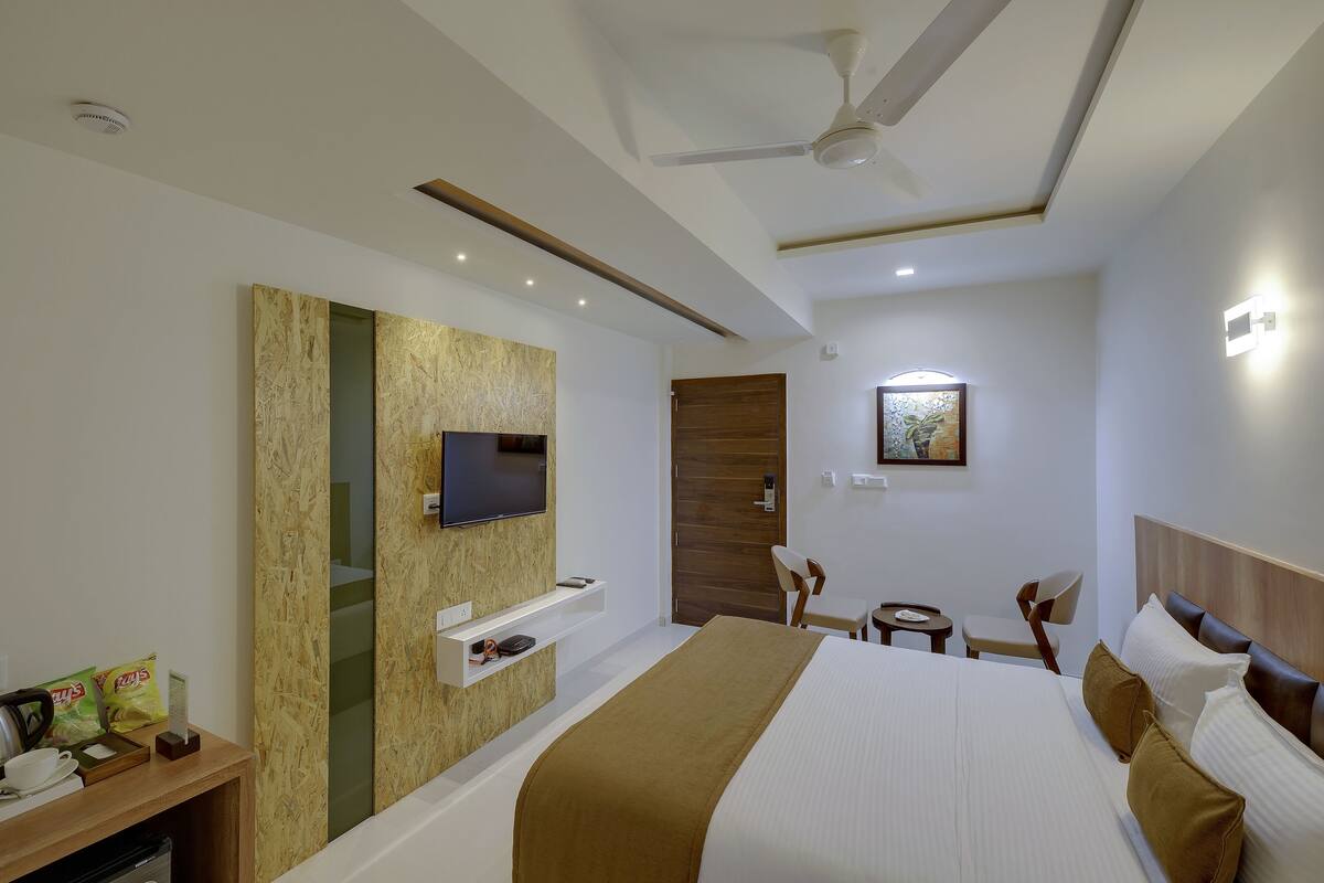 Quarto de hotel moderno com uma cama grande, televisão de tela plana na parede, uma pequena mesa com cadeiras e um ventilador de teto.