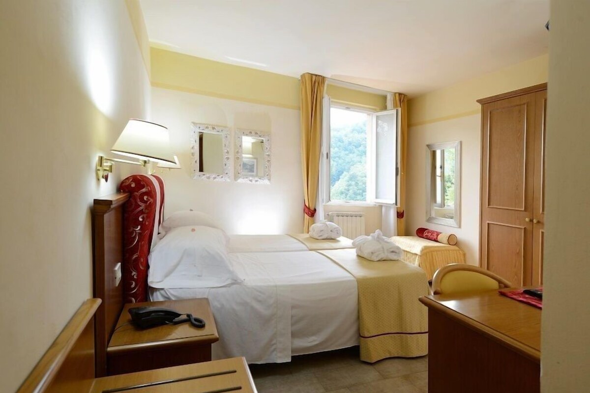 Quarto de hotel com duas camas, uma escrivaninha, uma cadeira, abajur, janela com cortinas e guarda-roupa.