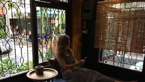 Mulher sentada num banco perto de uma janela com grades, segurando um telefone.