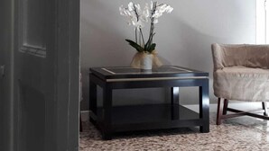 Um quarto com porta, espelho fixado na parede, pequena mesa com um vaso de flores e uma poltrona bege.