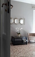 Um quarto com porta, espelho fixado na parede, pequena mesa com um vaso de flores e uma poltrona bege.