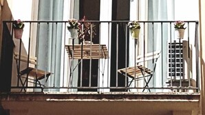 Varanda com balaustrada branca, mesa de madeira, duas cadeiras e plantas em vasos.