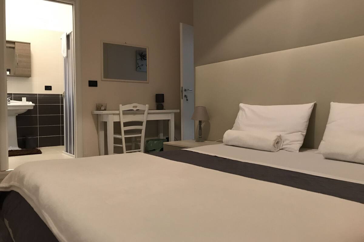 Quarto de hotel com cama, escrivaninha, cadeira, abajur e banheiro visível por uma porta aberta.