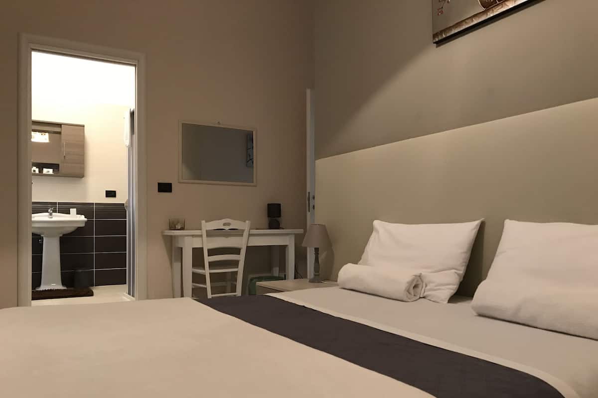 Quarto de hotel com cama, escrivaninha, cadeira, espelho e banheiro visível por uma porta aberta.