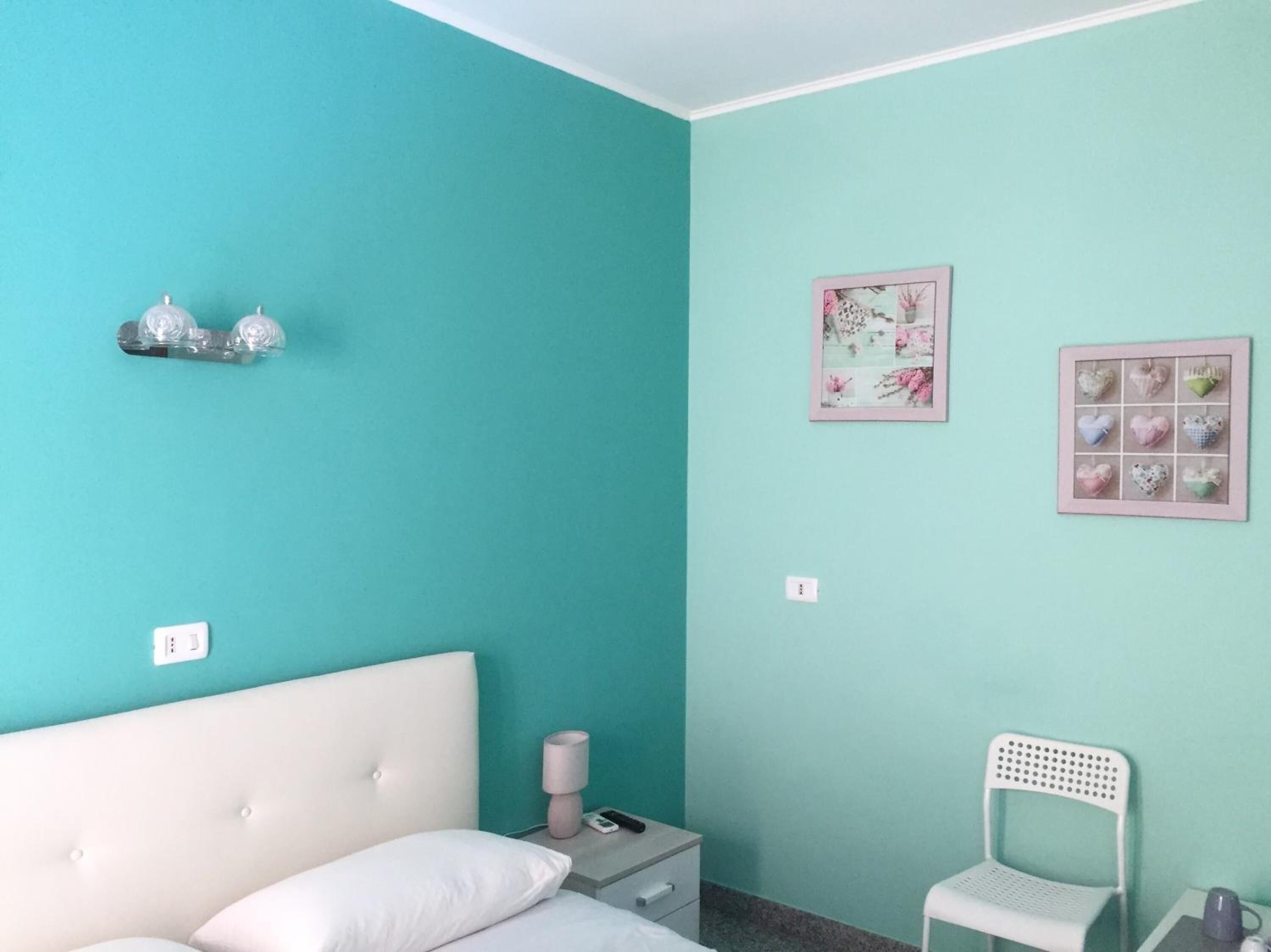 Quarto com paredes verde-água, uma cama branca, uma mesa de cabeceira com abajur e uma cadeira.