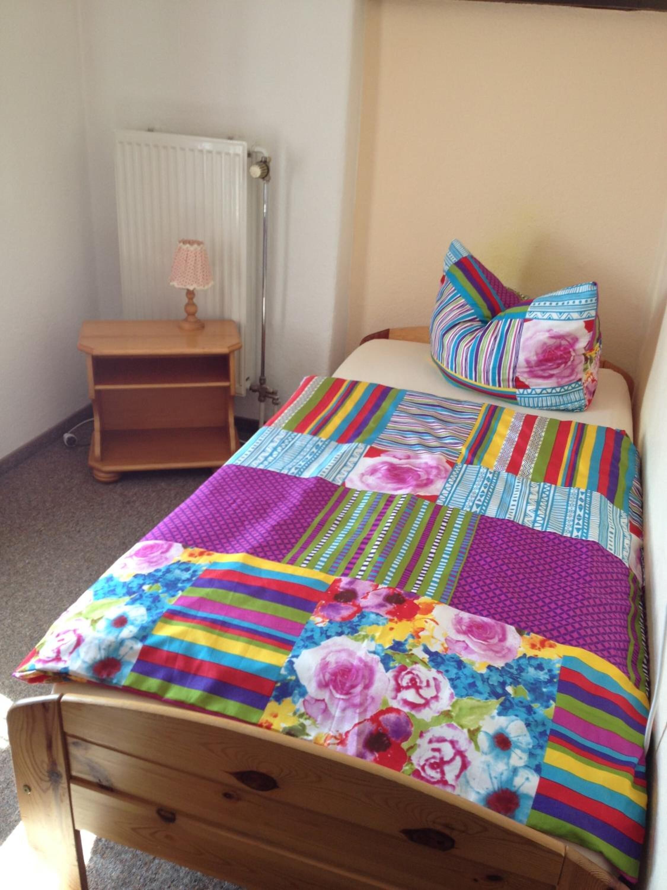 Uma cama de solteiro com um edredom colorido e almofada listrada, uma mesa de cabeceira de madeira com abajur e um radiador em um quarto com paredes simples.