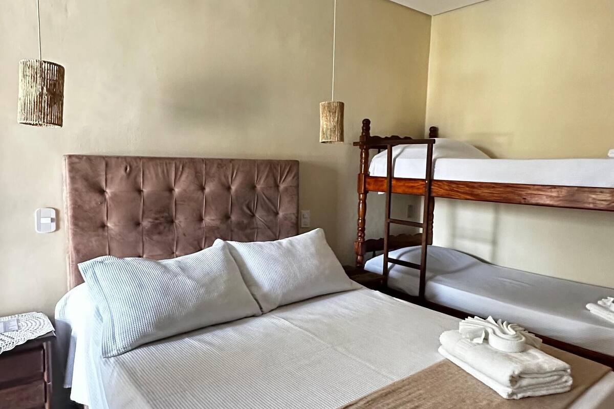 Quarto de hotel com uma cama grande, uma beliche de madeira, uma mesinha de cabeceira e um candeeiro suspenso.